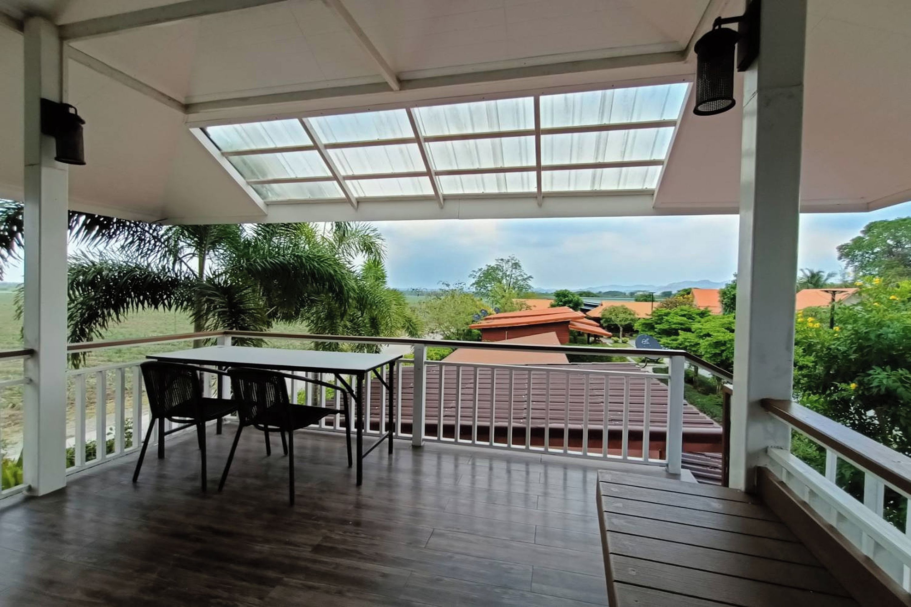 Uma área externa coberta com mesa e cadeiras, vista para vegetação e edifícios ao longe, e um corrimão de metal.