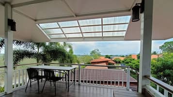 Uma área externa coberta com mesa e cadeiras, vista para vegetação e edifícios ao longe, e um corrimão de metal.
