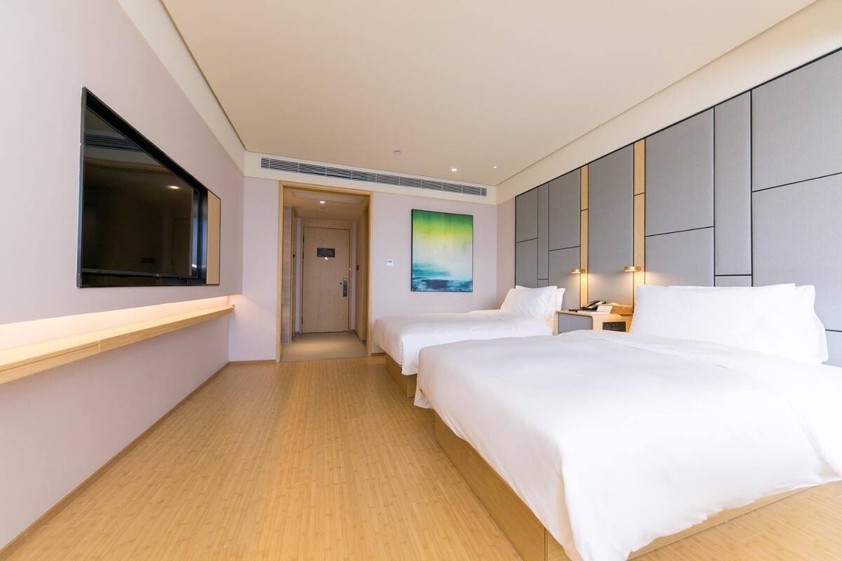 Quarto de hotel com duas camas, uma janela grande, um quadro e um corredor que leva a uma porta.