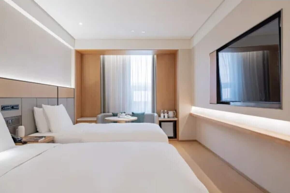 Quarto de hotel moderno com duas camas, uma mesa redonda pequena, televisão e uma janela grande com cortinas.