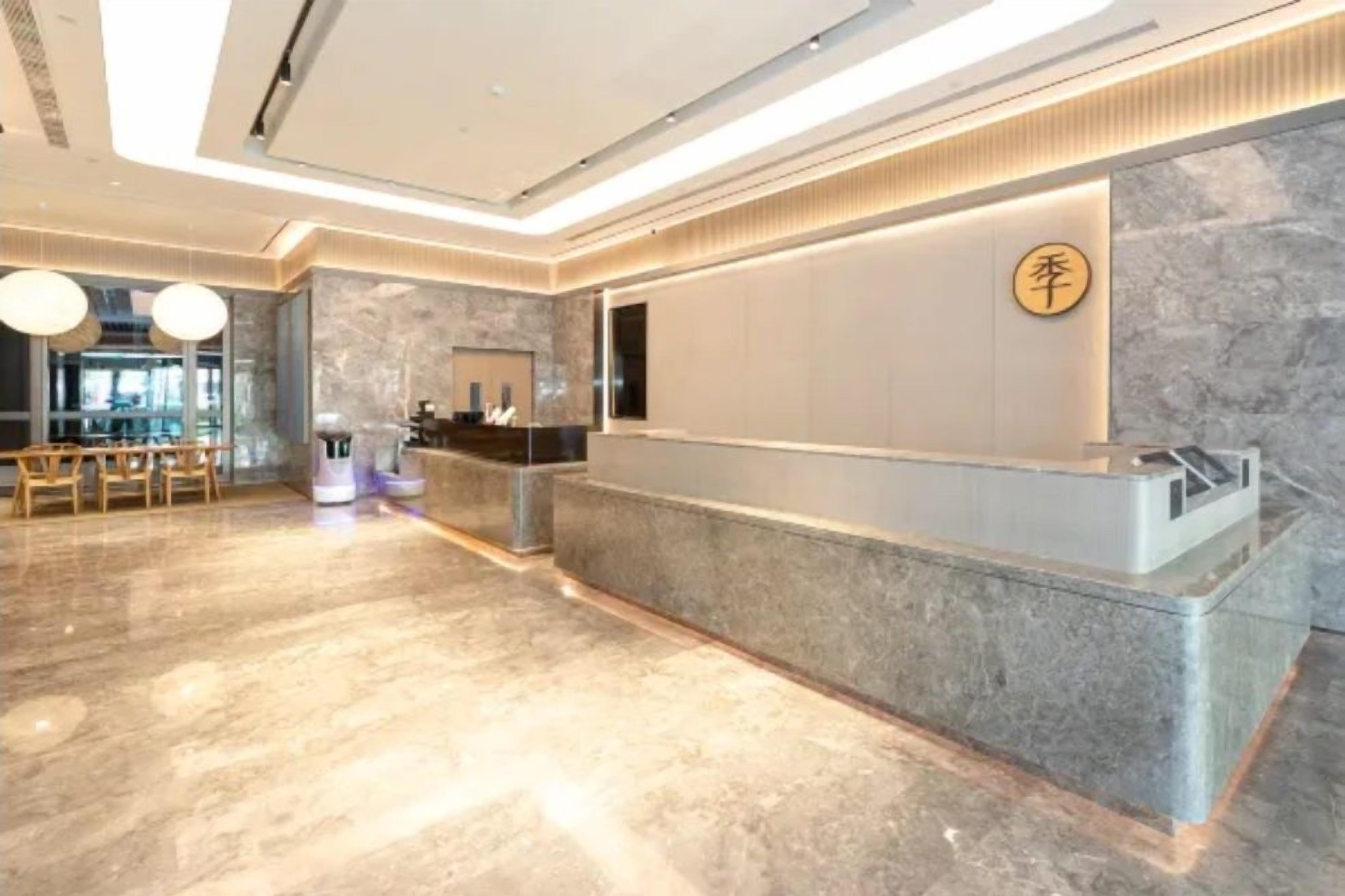 Um saguão de hotel moderno, com balcão de recepção, piso de mármore e área de estar com cadeiras de madeira.