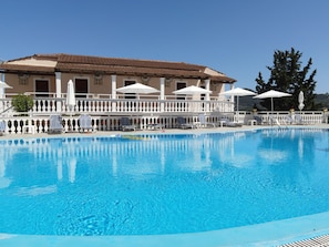 Piscina com espreguiçadeiras e guarda-sóis, um edifício com telhado de azulejos e uma cerca branca.