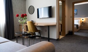 Quarto de hotel com cama, televisão na parede, pequena mesa com cadeiras, vaso com flores e escrivaninha de madeira.