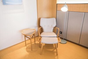 Um quarto com piso de madeira, uma cadeira branca com assento acolchoado, uma mesinha redonda pequena e um abajur.