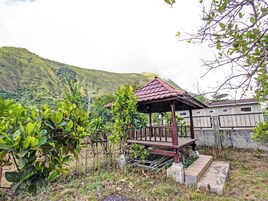Um gazebo de madeira com telhado de azulejos, rodeado por vegetação exuberante e uma encosta ao fundo.