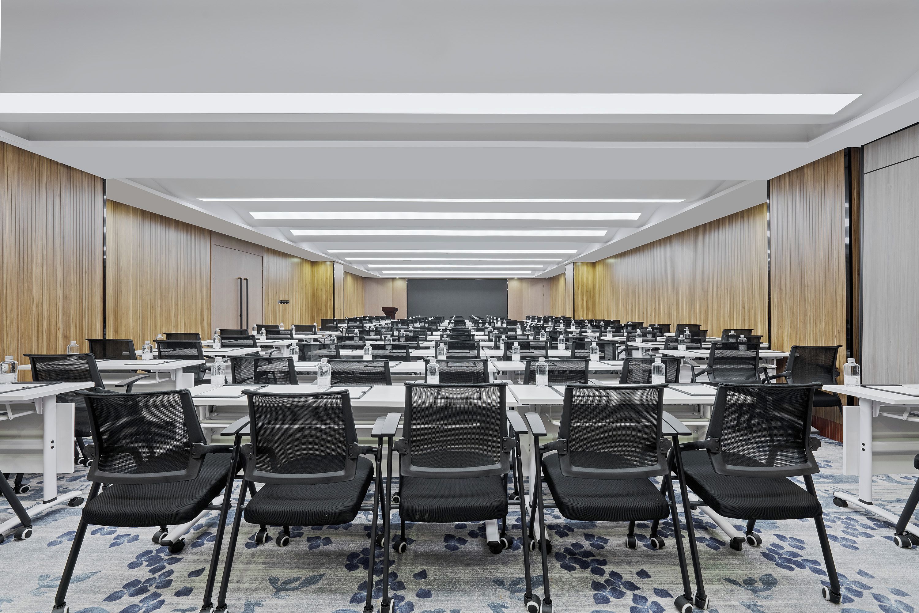 Uma sala de conferências com fileiras de mesas e cadeiras, equipada com computadores portáteis e garrafas de água.