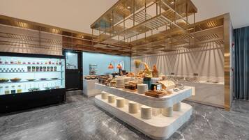 Um hall de hotel moderno com piso de mármore, uma vitrine de vidro com alimentos e um balcão central com pratos e tigelas.