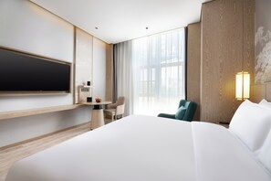 Quarto de hotel com uma cama grande, uma escrivaninha com cadeira, televisão e uma janela ampla.