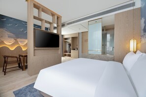 Quarto de hotel com uma cama grande, televisão de tela plana, móvel de madeira para televisão, televisão fixada na parede, espelho e banheiro visível através de uma porta aberta.