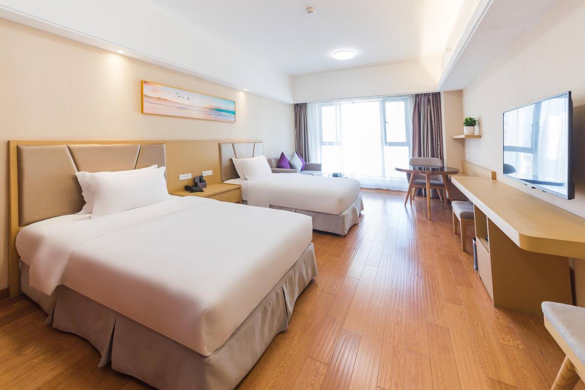 Quarto de hotel com duas camas, uma escrivaninha, televisão e uma janela grande.