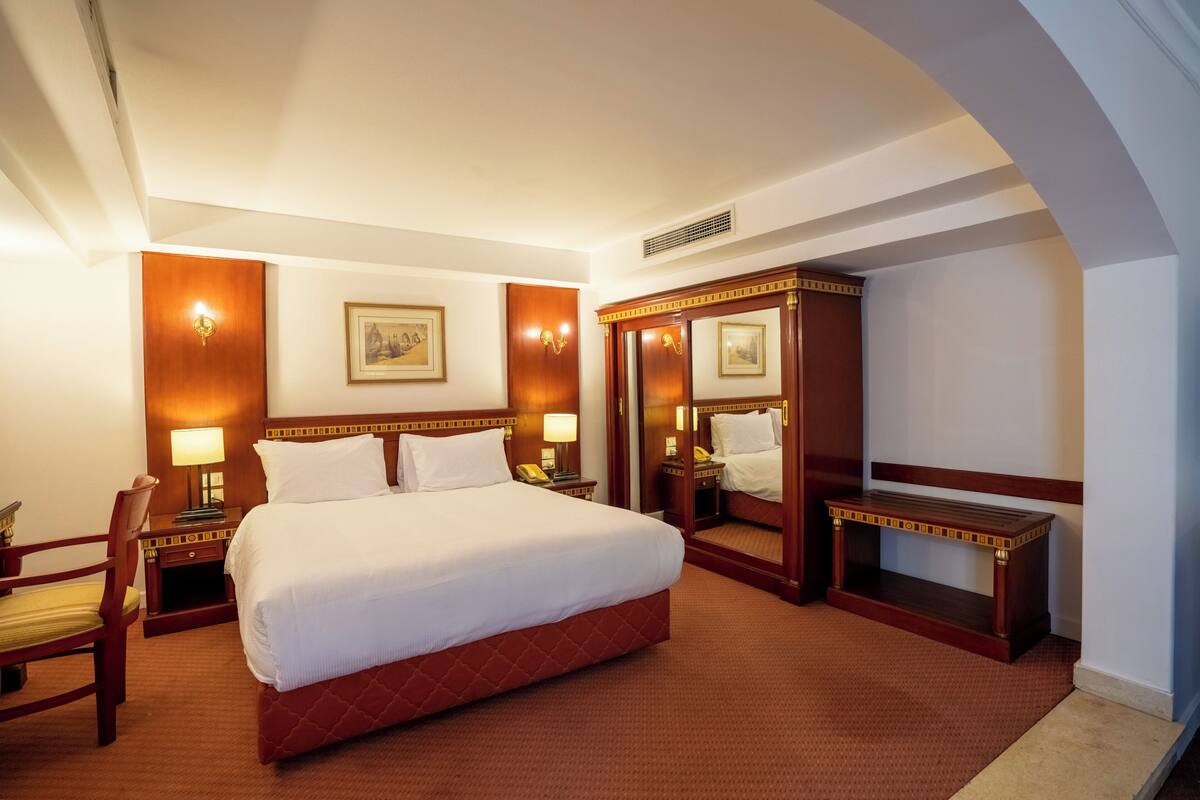 Quarto de hotel com uma cama grande, uma escrivaninha, uma cadeira, um espelho e uma mesinha.