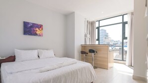 Quarto de hotel moderno com uma cama grande, uma escrivaninha com duas cadeiras, um quadro na parede e vista da cidade através de amplas janelas.
