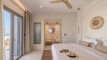 Quarto de hotel moderno com uma cama grande, um ventilador de teto, um espelho redondo e vista para a praia através de portas deslizantes.