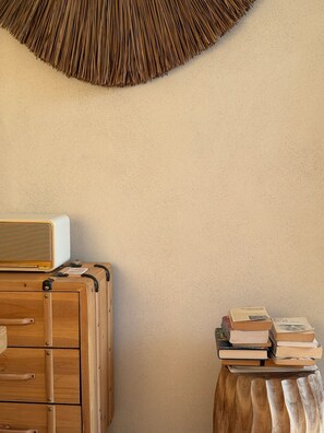 Uma cômoda de madeira com um rádio, um painel de parede tecido e uma pilha de livros sobre um banquinho de madeira.