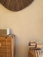 Uma cômoda de madeira com um rádio, um painel de parede tecido e uma pilha de livros sobre um banquinho de madeira.