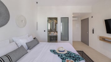 Quarto de hotel moderno com cama, um espelho grande, televisão e banheiro visível através de uma porta de vidro.