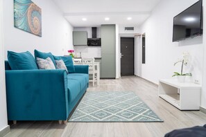 Uma sala de estar moderna com um sofá azul-turquesa, um tapete estampado, uma TV de tela plana e uma cozinha minimalista ao fundo.