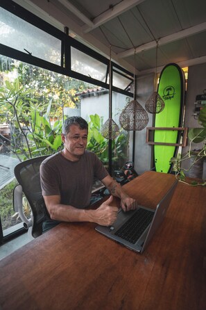 Um homem sentado à mesa de madeira com um laptop, uma prancha de surf e luminárias penduradas em um quarto com janelas amplas.