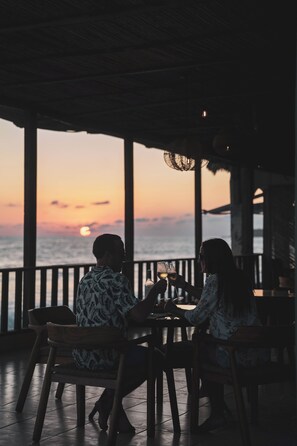 Um casal jantando em um restaurante à beira-mar, com vista para o pôr do sol no oceano.