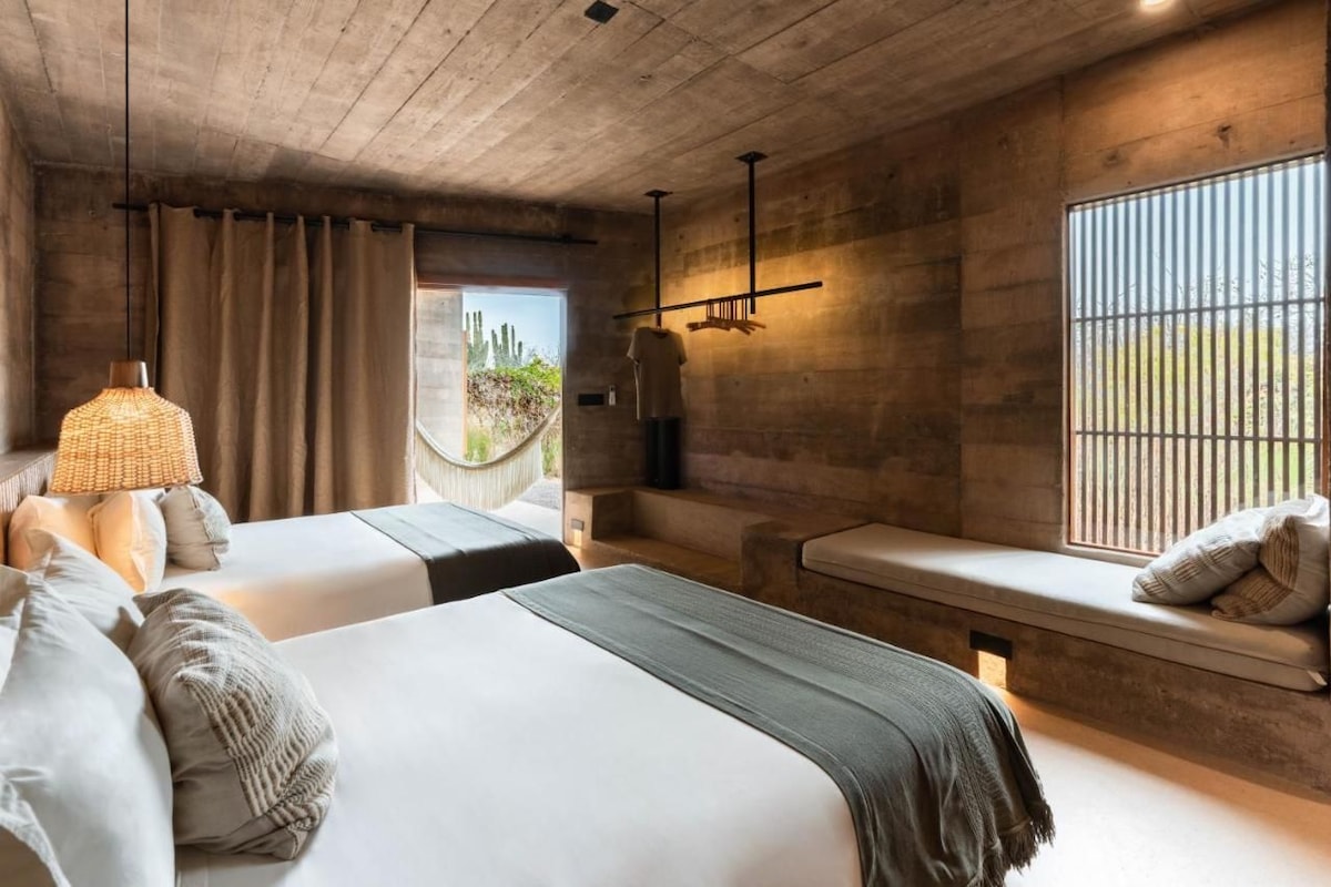 Quarto moderno com duas camas, um banco, uma luminária pendente e vista para o exterior.
