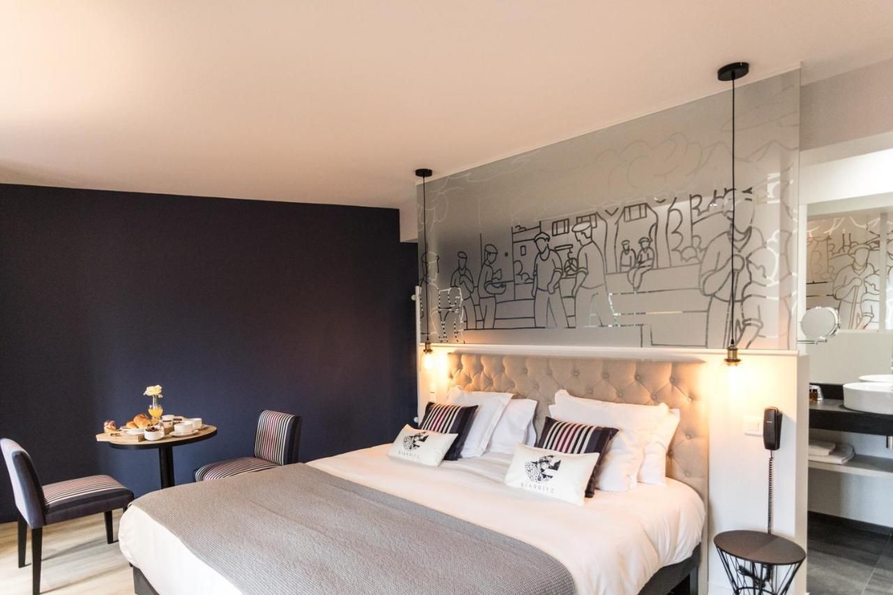 Quarto de hotel moderno com uma cama grande, uma mesa de cabeceira com abajur, uma cadeira e uma obra de arte na parede.