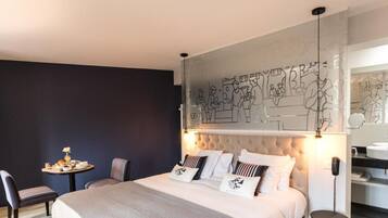 Quarto de hotel moderno com uma cama grande, uma mesa de cabeceira com abajur, uma cadeira e uma obra de arte na parede.
