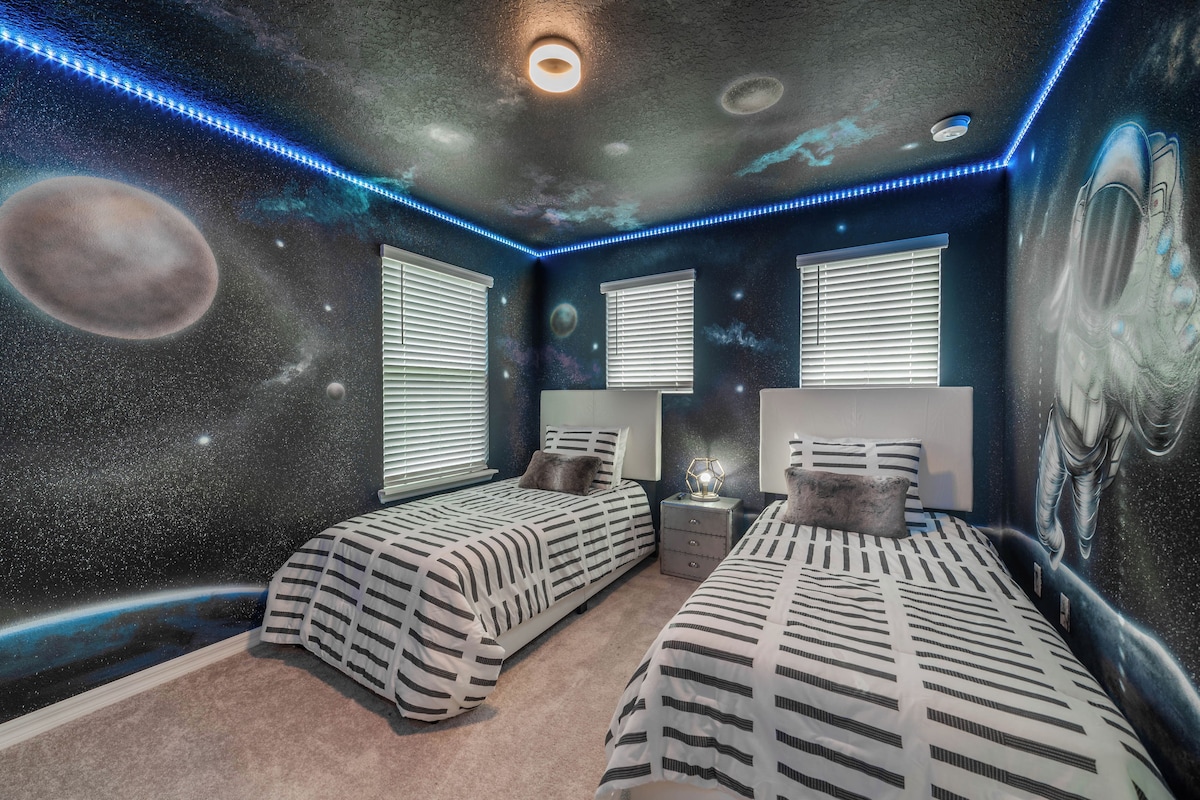 Quarto com duas camas, uma mesa de cabeceira, um mural na parede com tema espacial e um lustre.