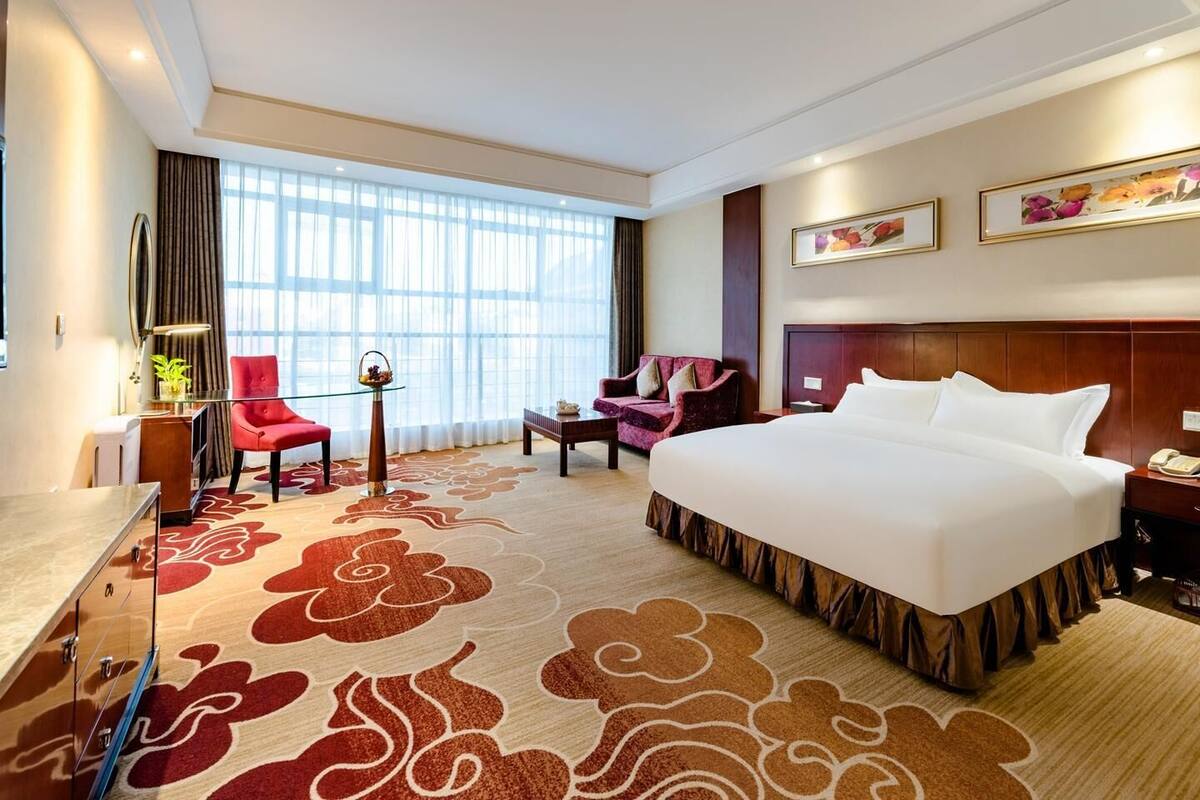 Quarto de hotel com uma cama grande, uma escrivaninha com cadeira, um sofá, uma mesa de centro e uma janela ampla.