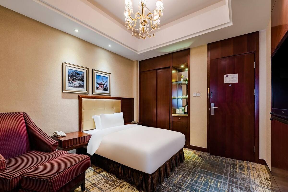 Quarto de hotel com cama grande, poltrona vermelha estofada, mesa de cabeceira de madeira, lustre e guarda-roupa.