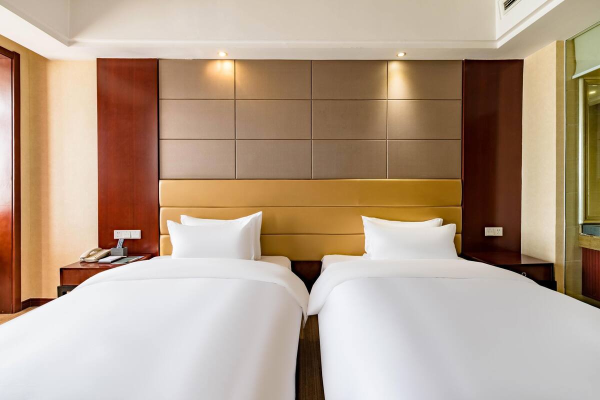 Quarto de hotel com duas camas, cabeceira de madeira, mesa de cabeceira com telefone e luminária fixada na parede.
