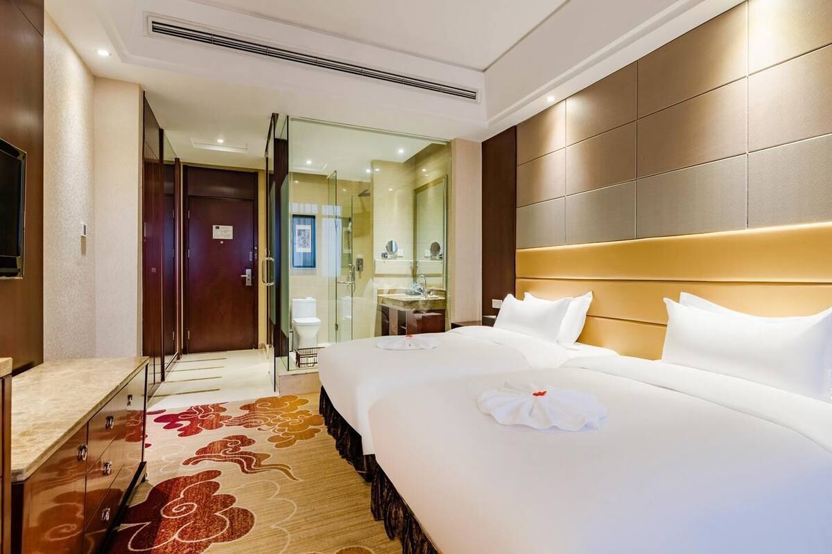 Quarto de hotel com uma cama grande, uma escrivaninha, um espelho e um banheiro visível através de uma porta de vidro.