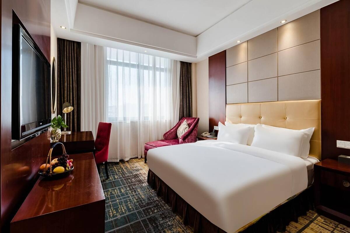 Quarto de hotel com uma cama grande, uma escrivaninha com televisão, uma cadeira e uma janela com cortinas.