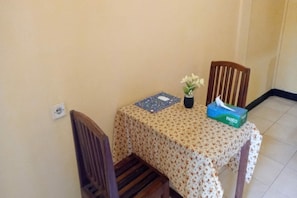 Uma pequena mesa de jantar com toalha florida, duas cadeiras de madeira, um vaso com uma única flor e uma caixa de lenços.