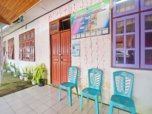 Um edifício com chão ladrilhado, teto de madeira e uma porta. Há plantas em vasos e cadeiras de plástico na parte externa.