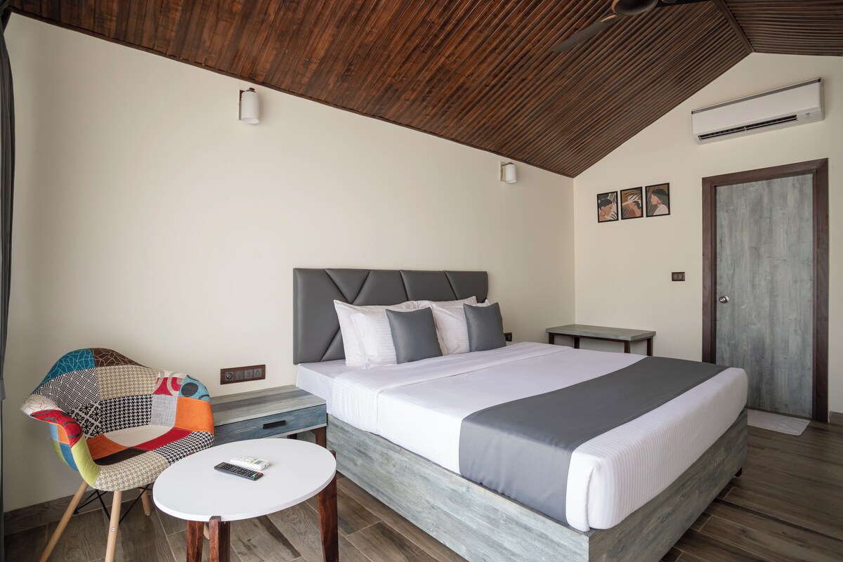 Quarto de hotel moderno com uma cama grande, uma cadeira colorida, uma mesa pequena e teto de madeira.