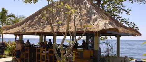 Um bar ao ar livre com telhado de palha, mesas e cadeiras, com vista para o mar.