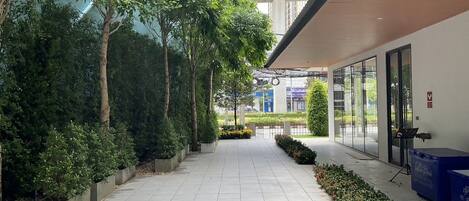 Uma galeria coberta com vasos de plantas e árvores, que leva a um edifício com portas de vidro.