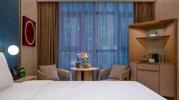 Quarto de hotel com cama, duas cadeiras, uma mesinha com um vaso de flores, um minibar e uma janela grande com cortinas.