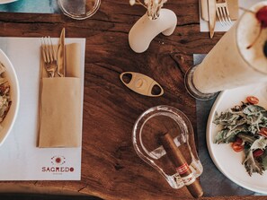 Uma mesa de madeira com uma salada numa placa branca, um copo com bebida e um guardanapo com talheres.