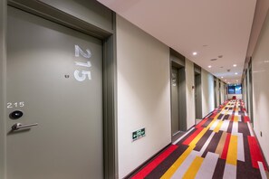 Corredor de hotel com carpete estampado, uma porta com a inscrição '215' e um sinal de saída de emergência.