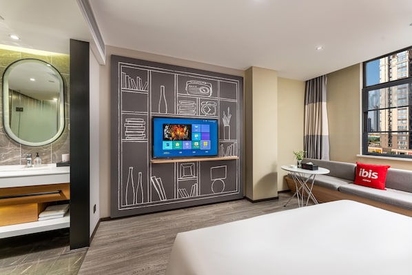 Quarto de hotel moderno com uma cama grande, uma televisão na parede, uma mesa redonda pequena e um sofá com um travesseiro vermelho.