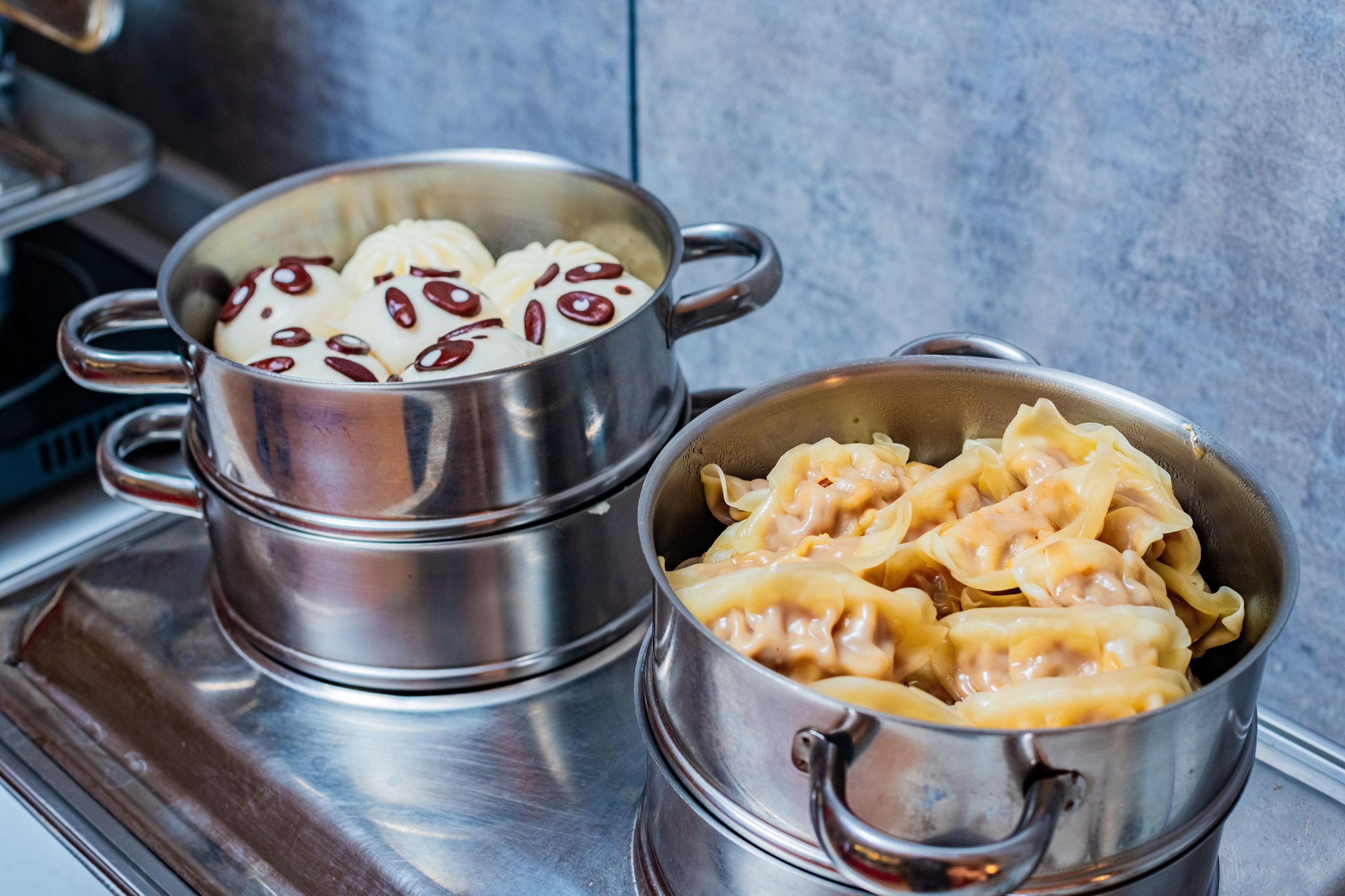 Duas panelas de aço inoxidável em um fogão, uma com dumplings e outra com uma substância branca e sementes vermelhas.