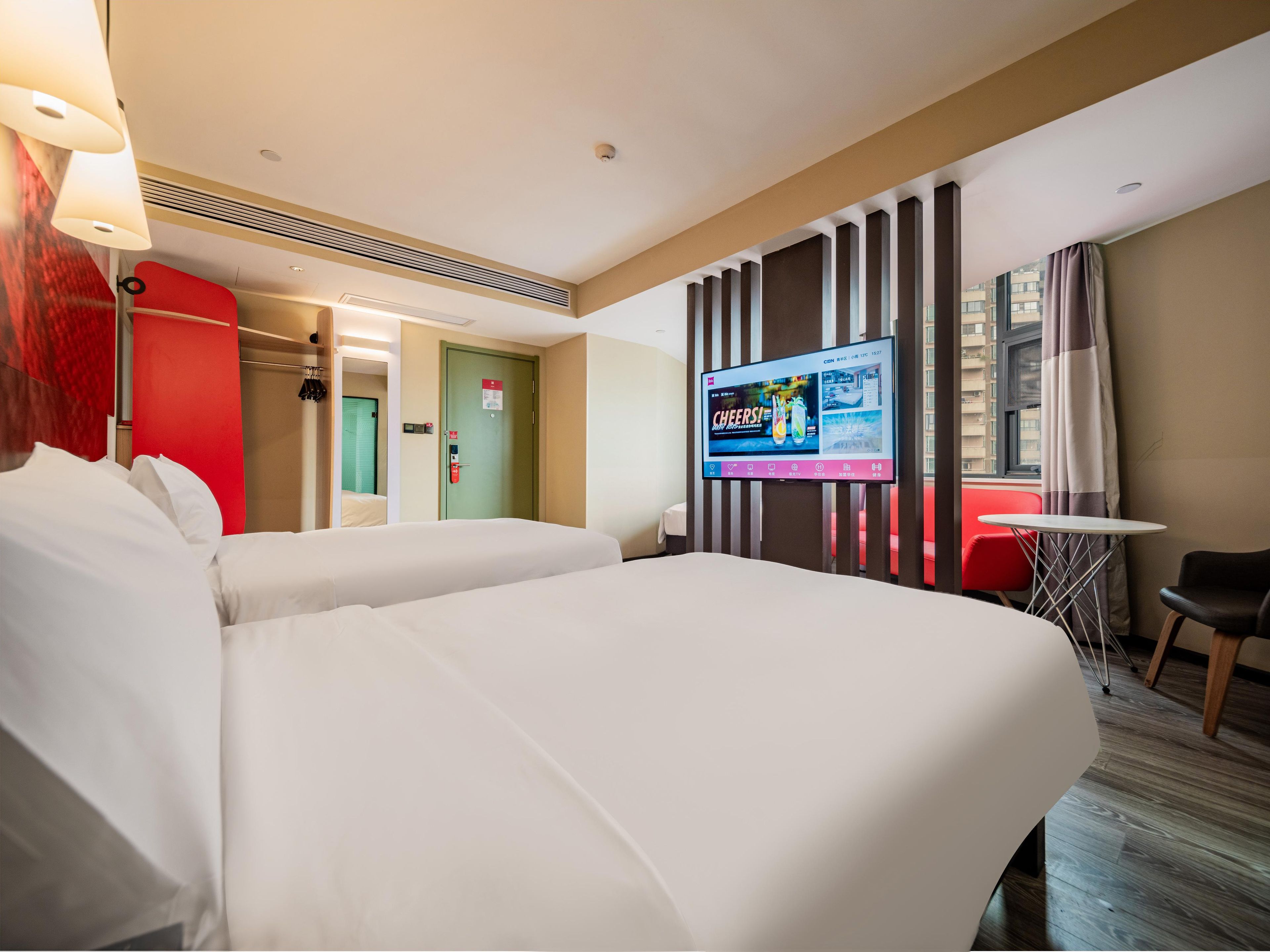 Quarto de hotel com duas camas, televisão na parede, uma mesinha e janela com cortinas.