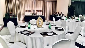 Uma sala de jantar de hotel com mesas redondas, cadeiras brancas e cortinas ao fundo.