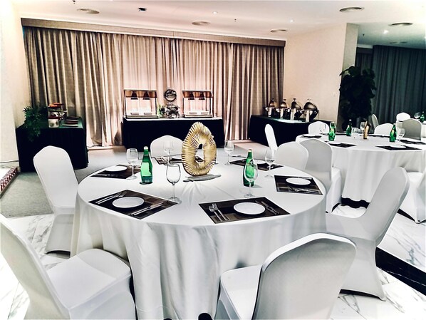 Uma sala de jantar de hotel com mesas redondas, cadeiras brancas e cortinas ao fundo.