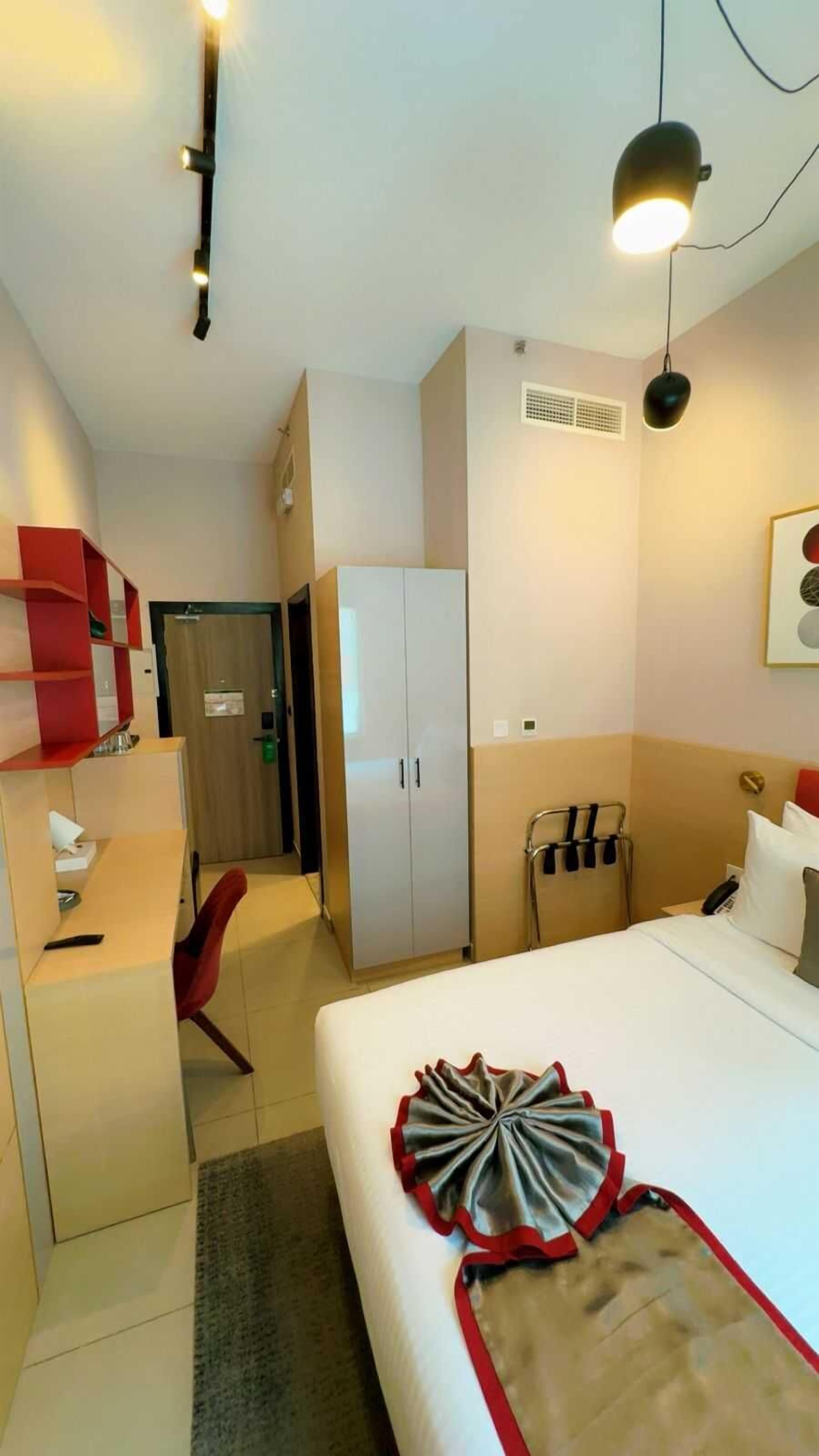 Quarto de hotel com cama, escrivaninha, cadeira, guarda-roupa e luminária pendente.