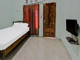 Uma cama de solteiro com roupa de cama branca, uma porta de madeira, uma televisão fixada na parede e um chão ladrilhado.