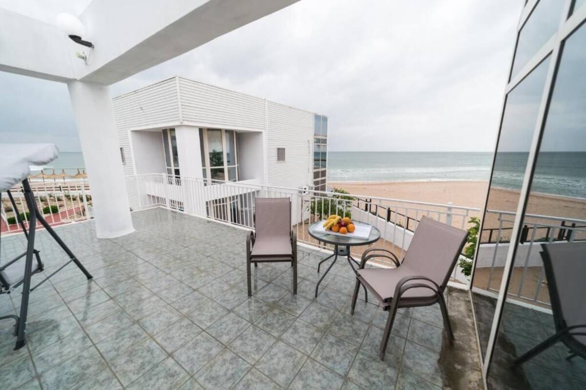 Uma varanda com mesa e cadeiras, um edifício branco e vista para a praia.