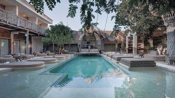 Uma piscina com espreguiçadeiras, uma estrutura com telhado de palha e um edifício moderno com varandas.