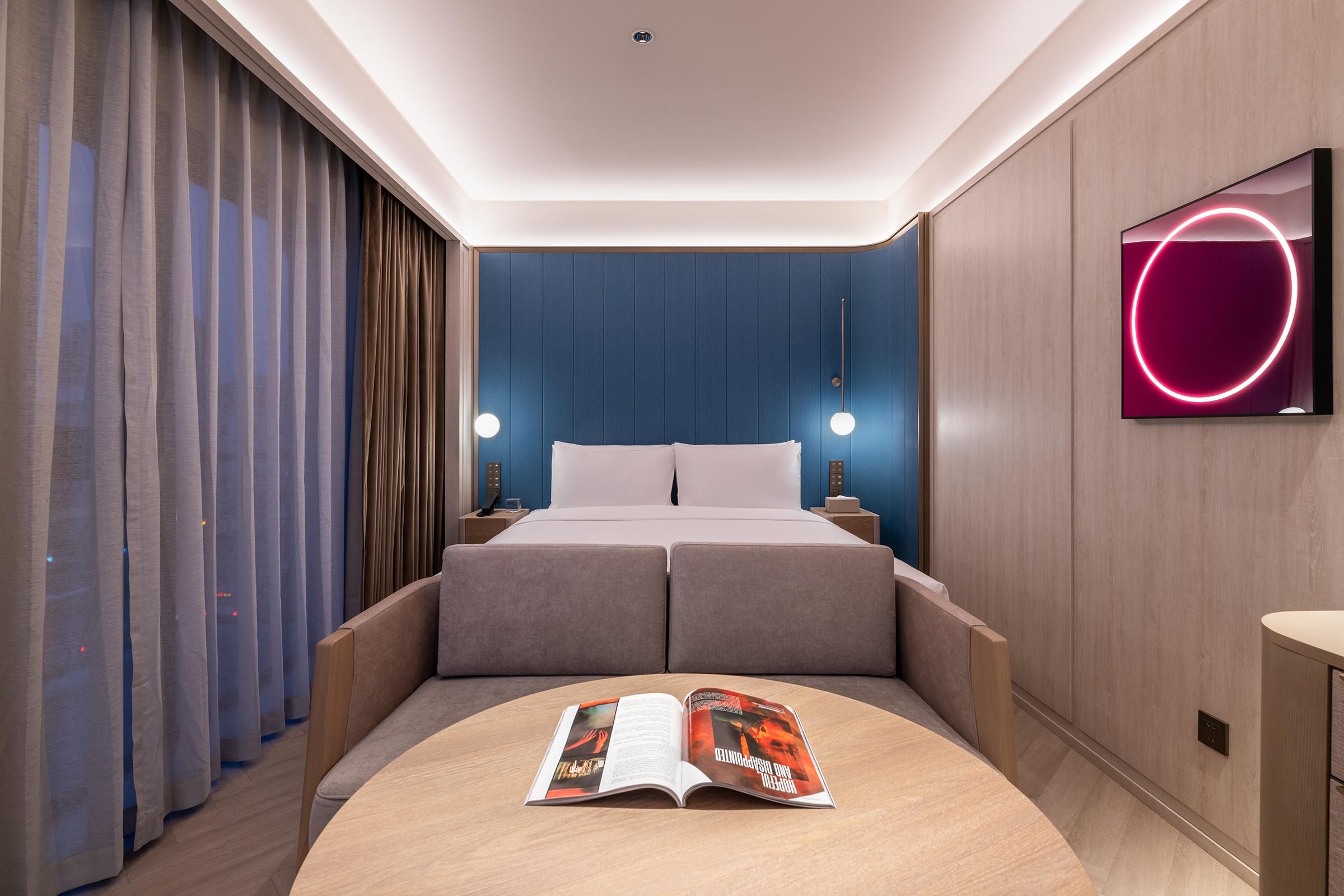 Quarto de hotel moderno com cama, mesas de cabeceira, uma mesa redonda com uma revista e um detalhe na parede com iluminação neon.